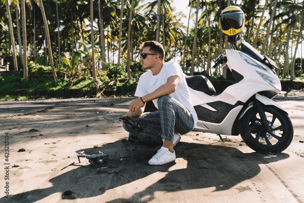 Dreamy young man squatting by motorcycle with drone controller on palms ...