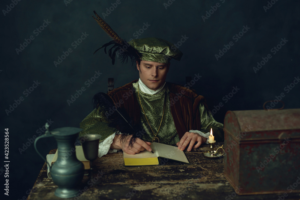 Renaissance man sits behind table writing in a notebook by candlelight in the evening. Stock ...