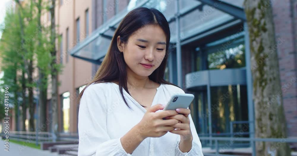 Smiling Asian woman engaged into online chatting, walking in urban city ...