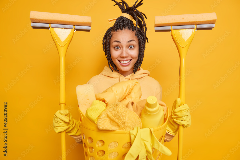 Positive ethnic woman holds two mops going to wash floor poses with ...