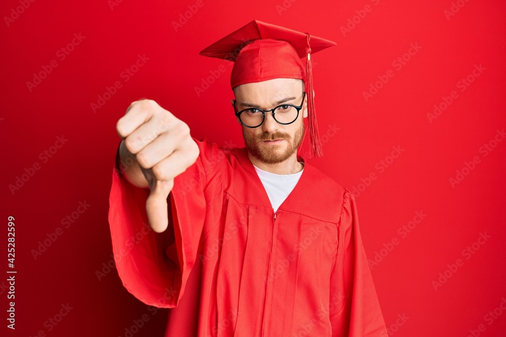 Young redhead man wearing red graduation cap and ceremony robe looking ...