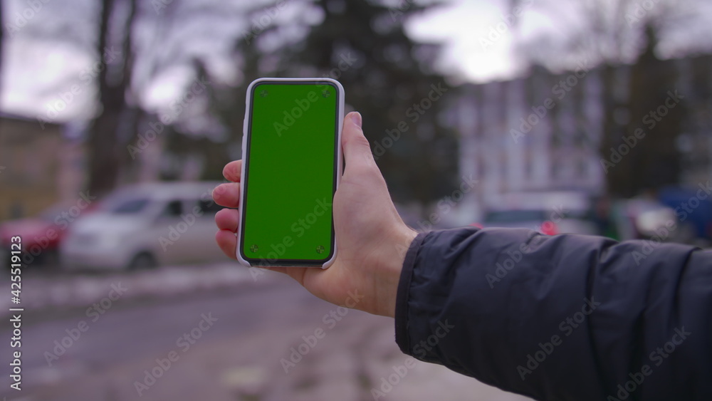 Hand man uses holding a mobile telephone with a vertical green screen background on of street ...