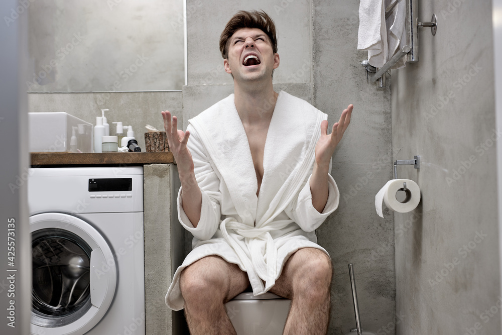 Man in bathrobe suffering from stomach ache on toilet bowl in bathroom ...