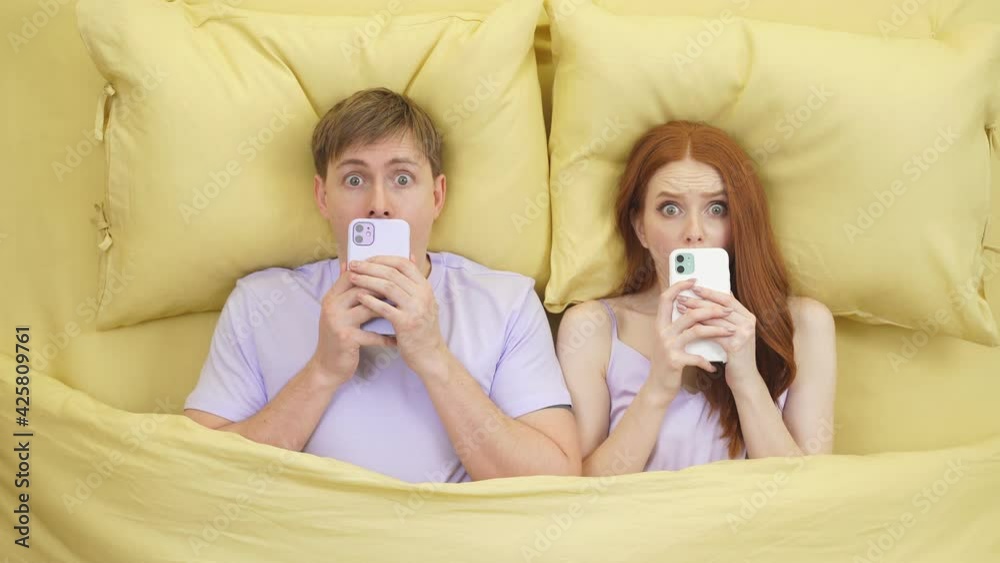 Couple in shock, lying in bed at night while using mobile phones, top ...
