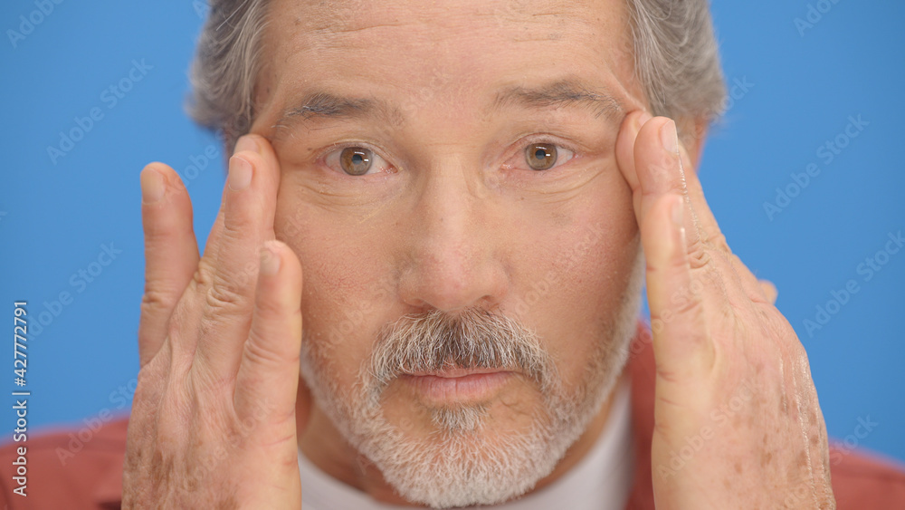 In front of the blue background, the old man looks at his wrinkles on ...