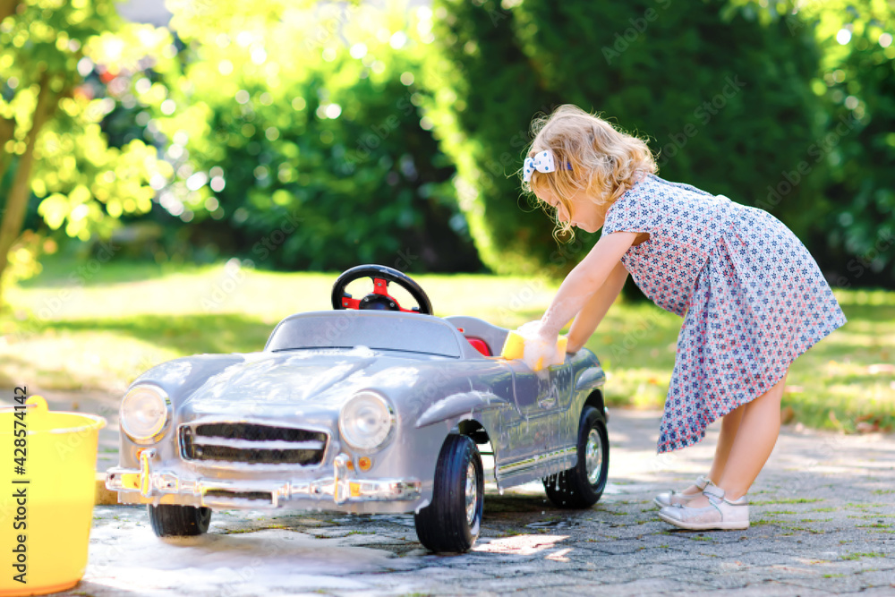 Cute gorgeous toddler girl washing big old toy car in summer garden ...