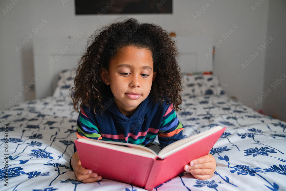 Afro little girl laying on bed with a red book reading front view Stock ...