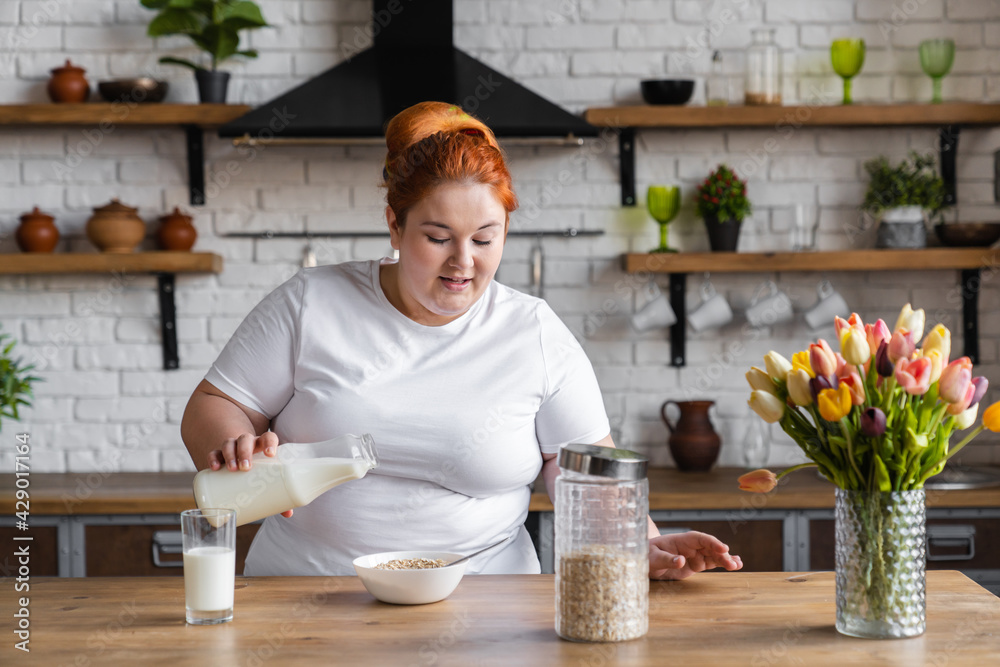 Young chubby woman standing in kitchen pouring milk into oat-flakes ...