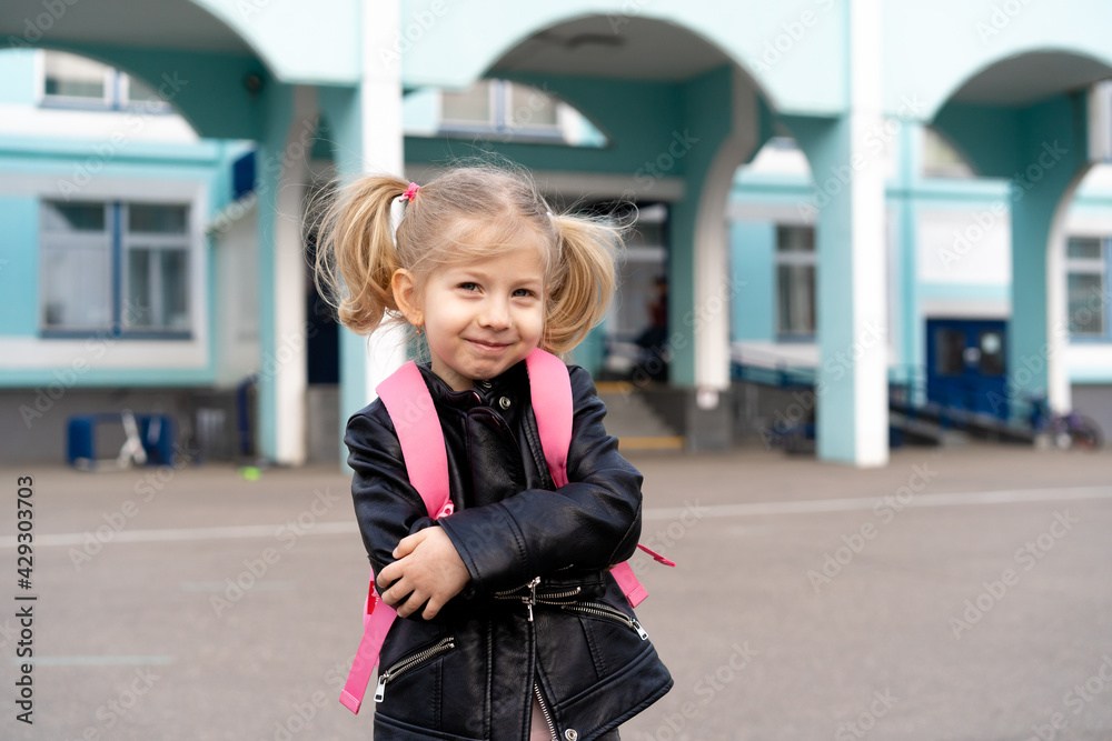 Happy little girl first-grader stands near school with a pink backpack ...