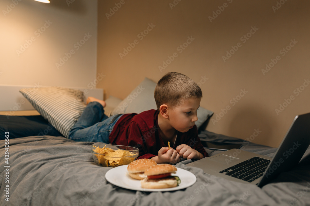 young boy eating unhealthy food and using laptop while sitting in his ...