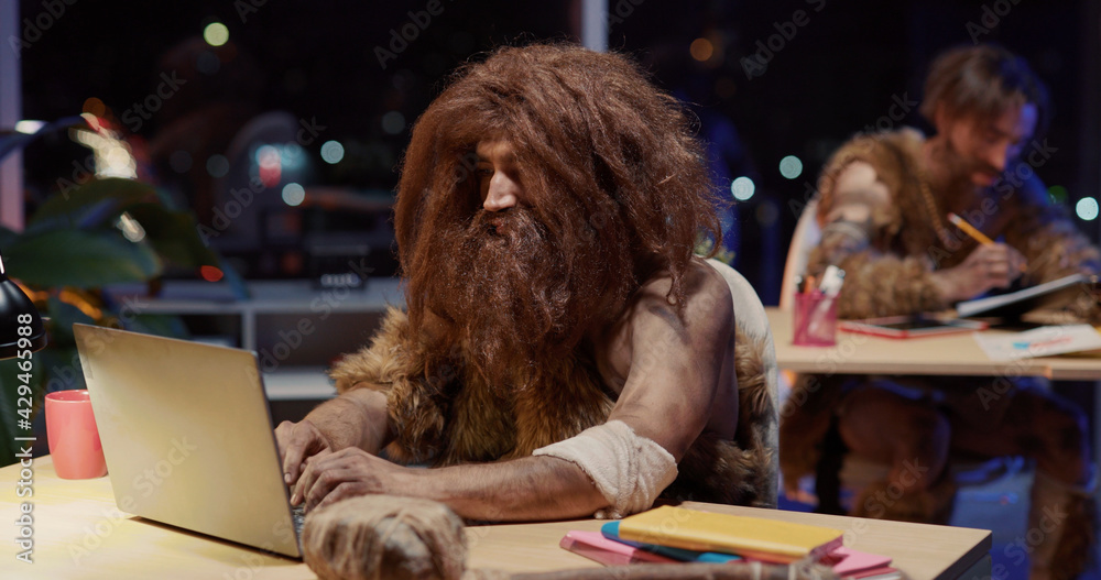 Portrait of confident savage man in cave animal clothes using laptop ...