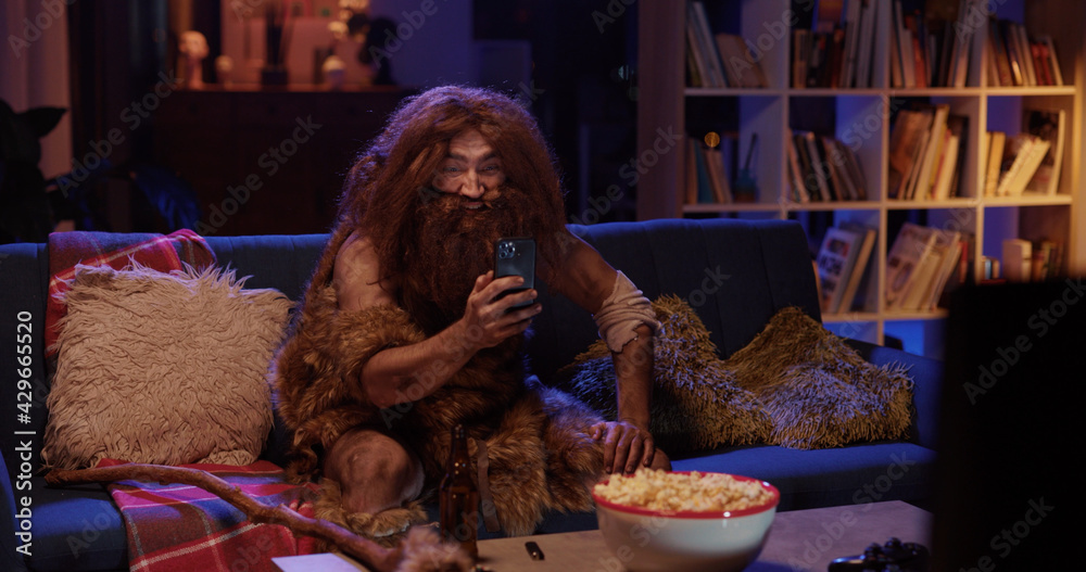 Furry bearded human ancestor in animal fur sitting on couch watching TV ...