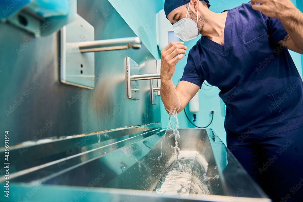 young Caucasian surgeon in scrubs washes his hands before entering the ...