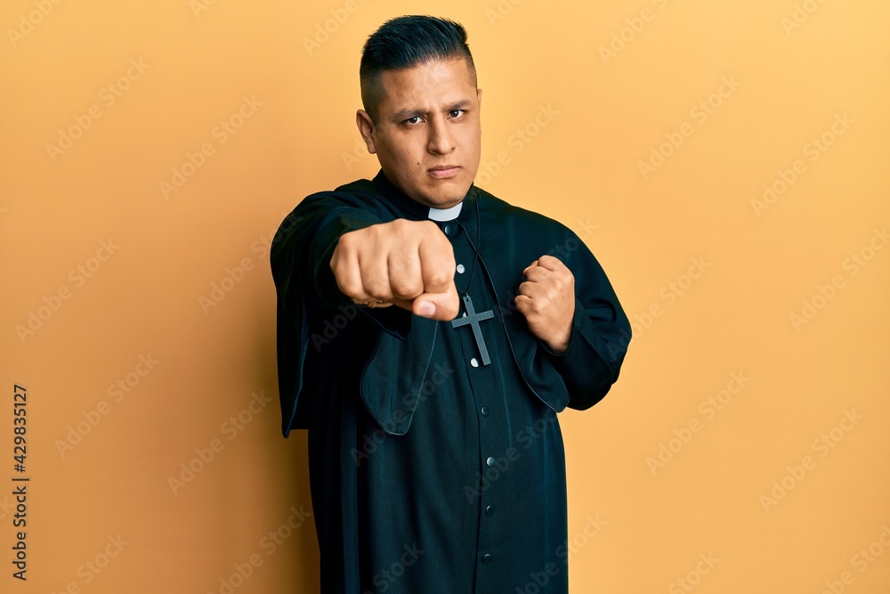 Young latin priest man standing over yellow background punching fist to ...