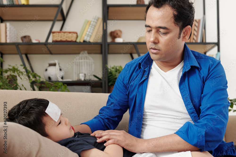 Serious worried father sitting next to is sick child and telling him ...