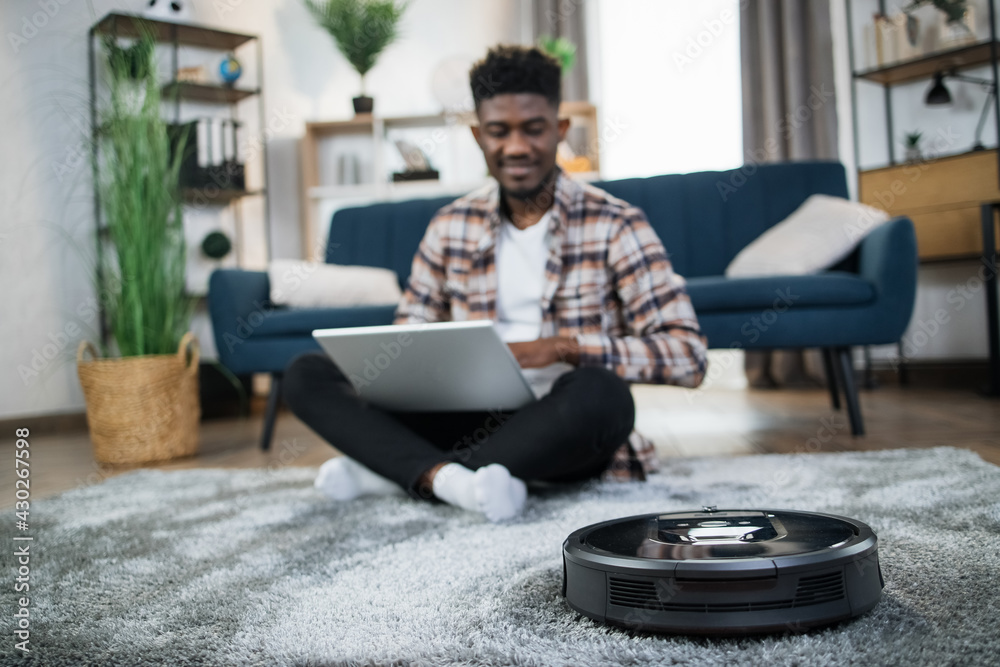 Positive african guy sitting on floor with laptop while robotic vacuum ...