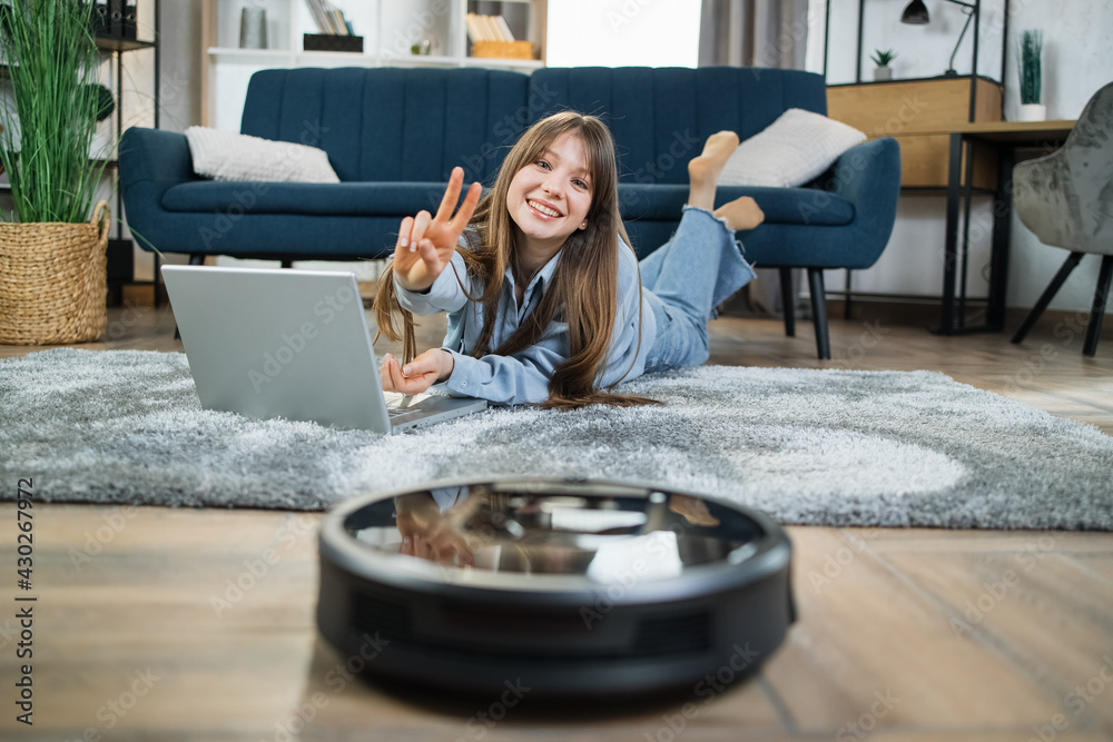 Cheerful young woman lying on carpet with wireless laptop and showing ...