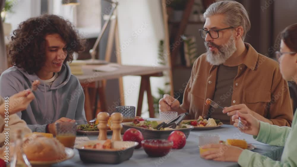 Mature Caucasian man eating food and speaking with cheerful teenage son ...