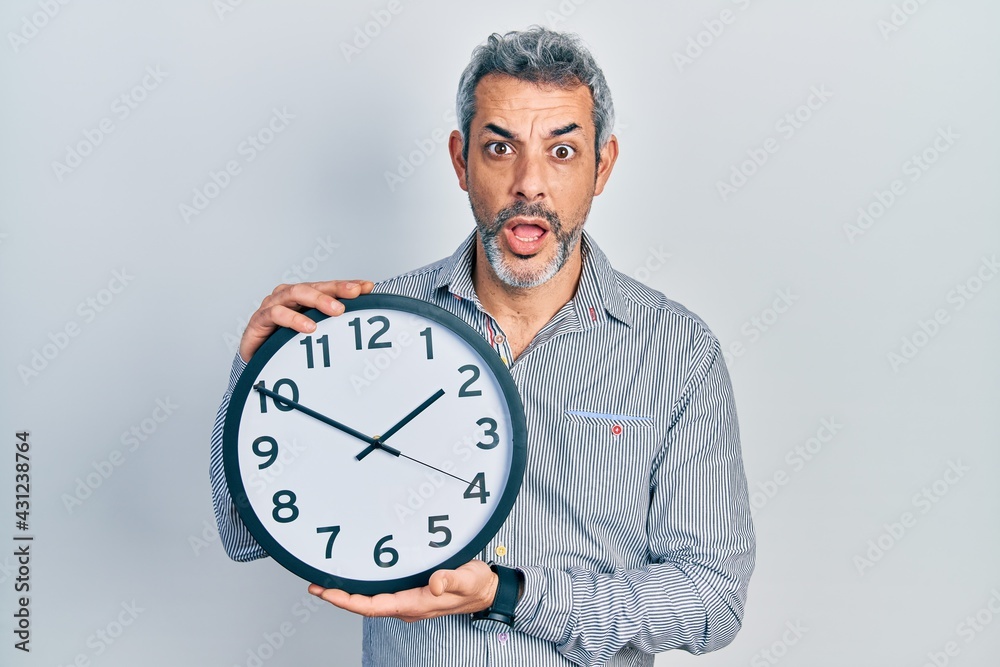 Handsome middle age man with grey hair holding big clock afraid and ...