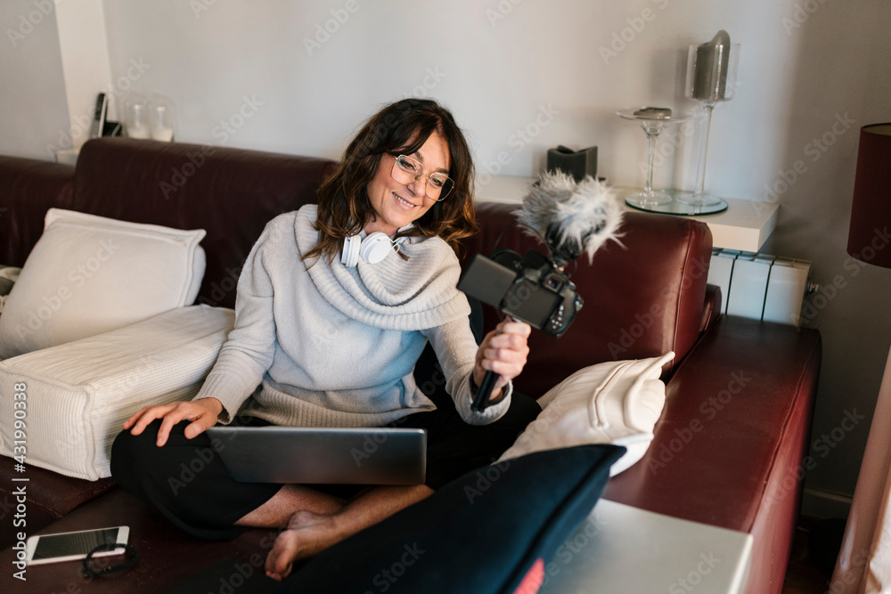 Smiling female vlogger with laptop using camera while vlogging at home ...