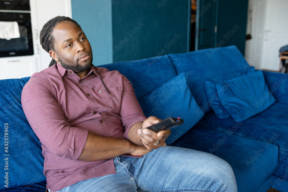 Bored African-American guy sitting on the couch at home, switching tv, sleepy and melancholy ...