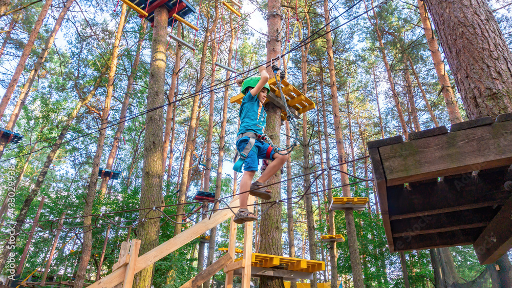 A boy in a helmet climbs a rope ladder stretched between trees in a ...