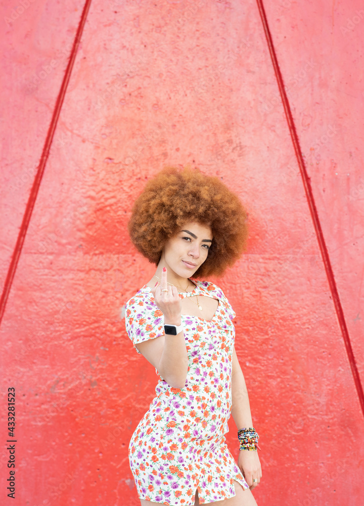 Young woman gesturing obscene hand sign while standing in front of red ...