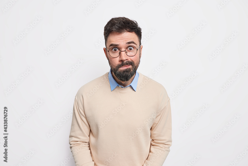 Puzzled bearded adult man purses lips reacts on astonishing news wears ...