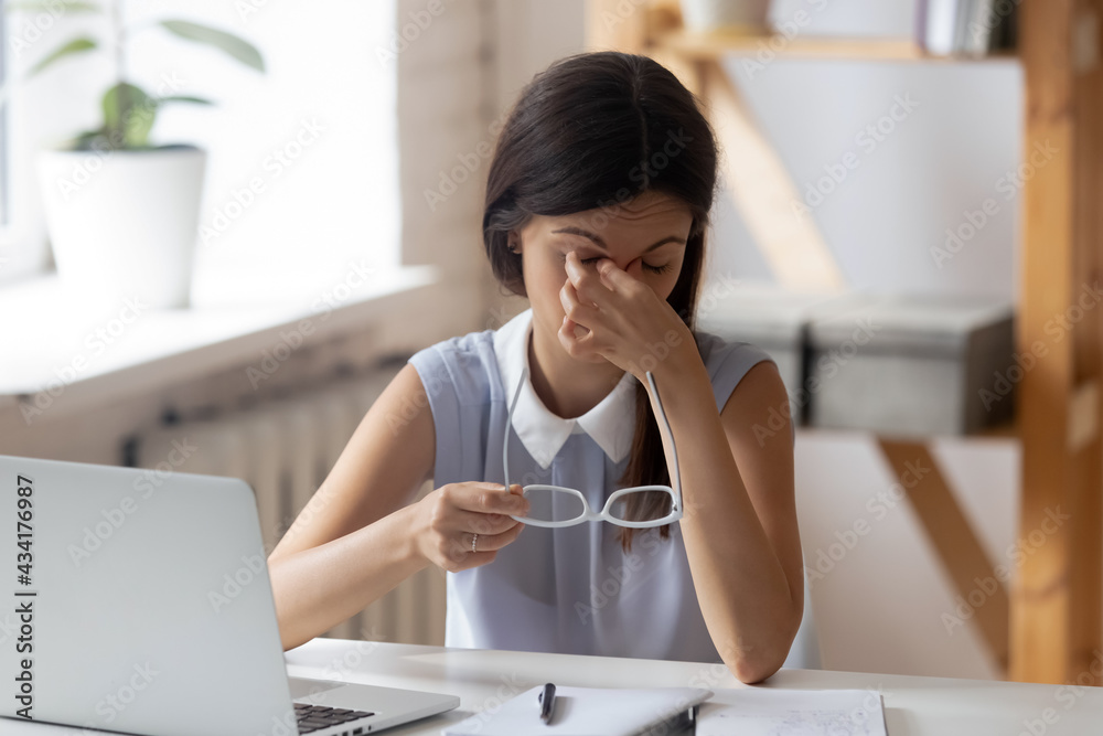 Exhausted young Caucasian woman work on laptop take off glasses suffer ...
