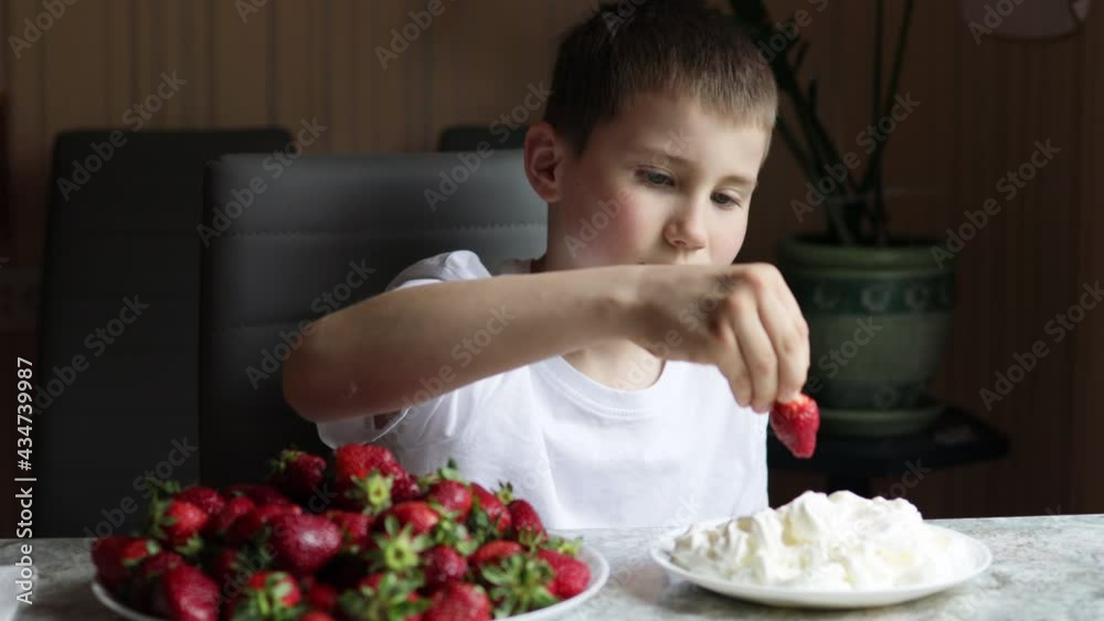 Boy eating strawberries with cream sitting near kitchen table at home ...