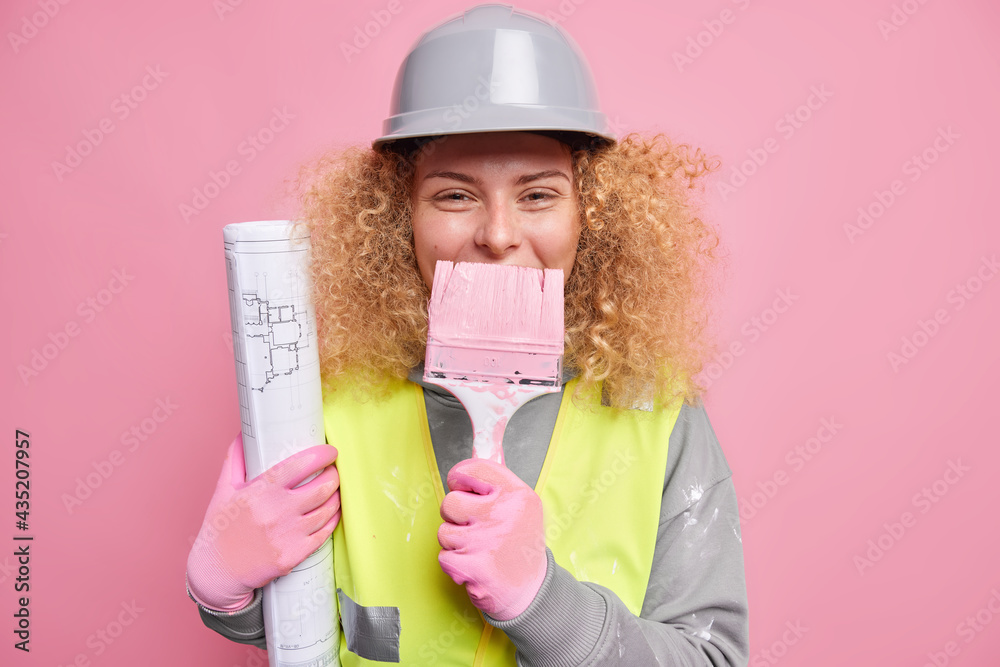 Positive female engineer with curly bushy hair keeps paint brush over ...