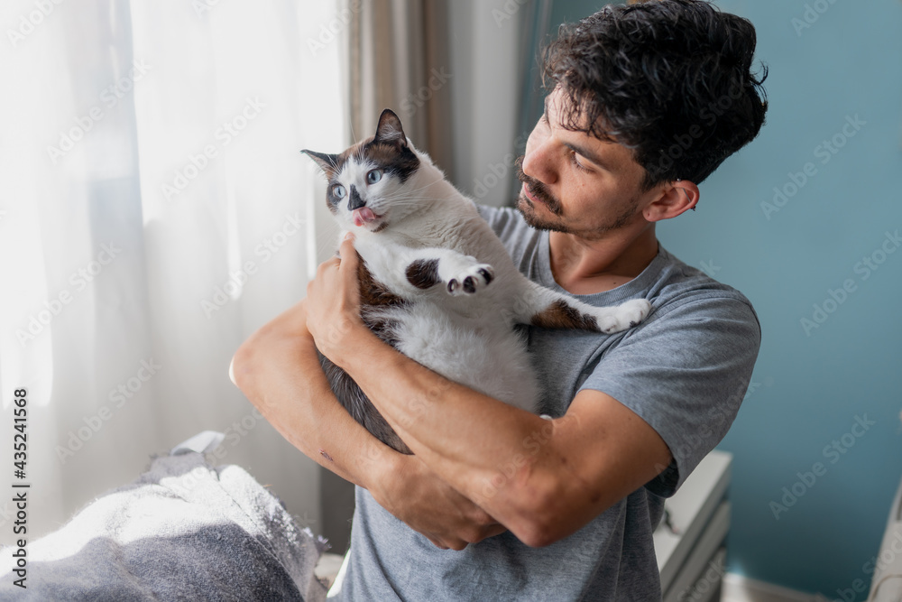 young man hugs a black and white cat with blue eyes in the living room ...