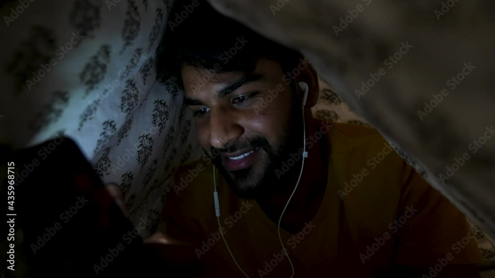 Indian man watching streaming video on a smart phone laying on the bed ...