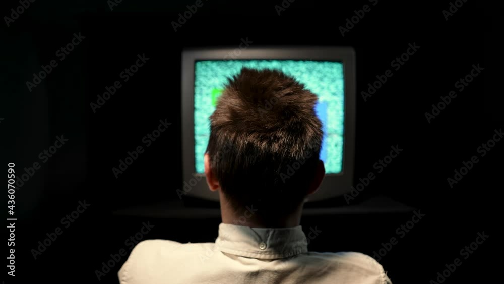 Rear view of a man sitting in front of an old TV in a dark studio on a ...