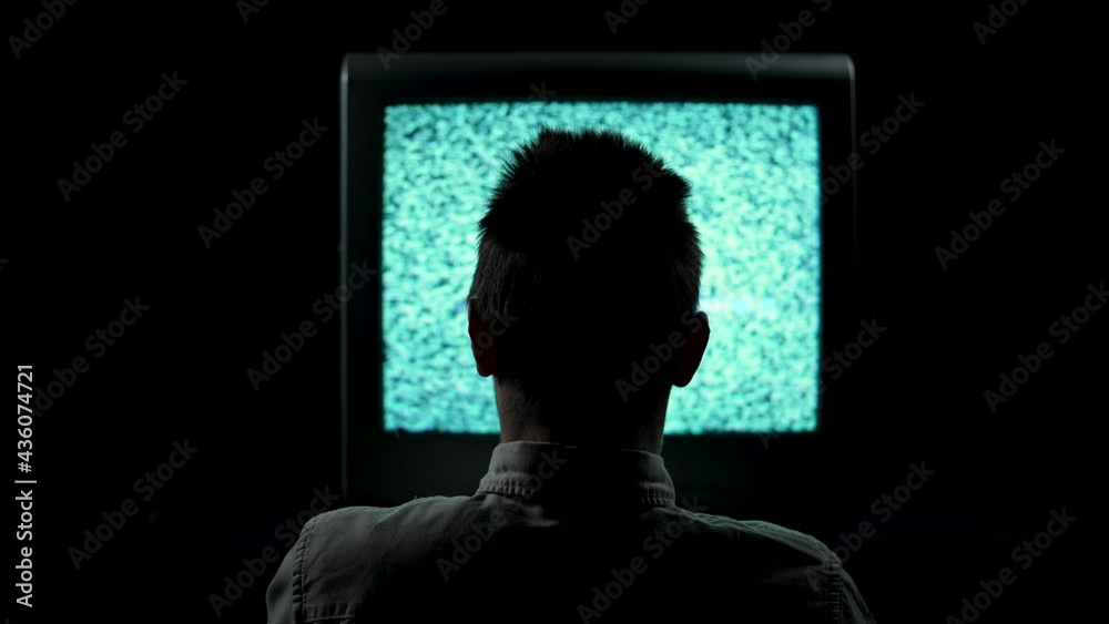 Rear view of a silhouette of a man sitting in front of a flickering TV ...