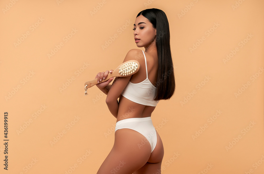 Young Asian woman in white lingerie makes anti-cellulite massage with a dry brush on a beige ...