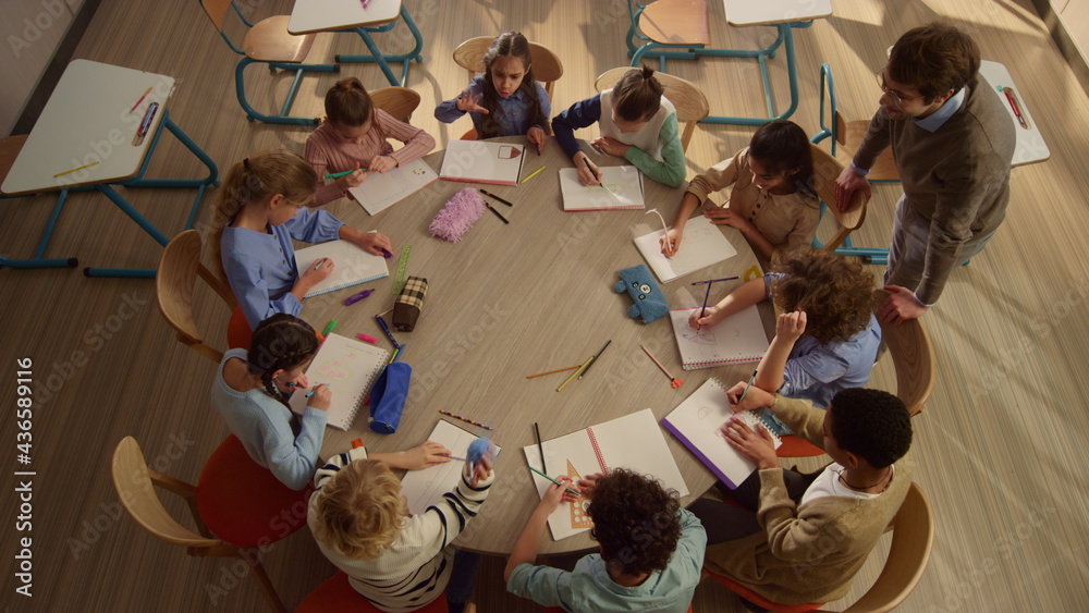 Schoolchildren having lesson at round table. Male teacher helping kids ...