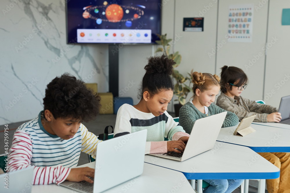 Diverse group of children sitting in row at school classroom and using ...