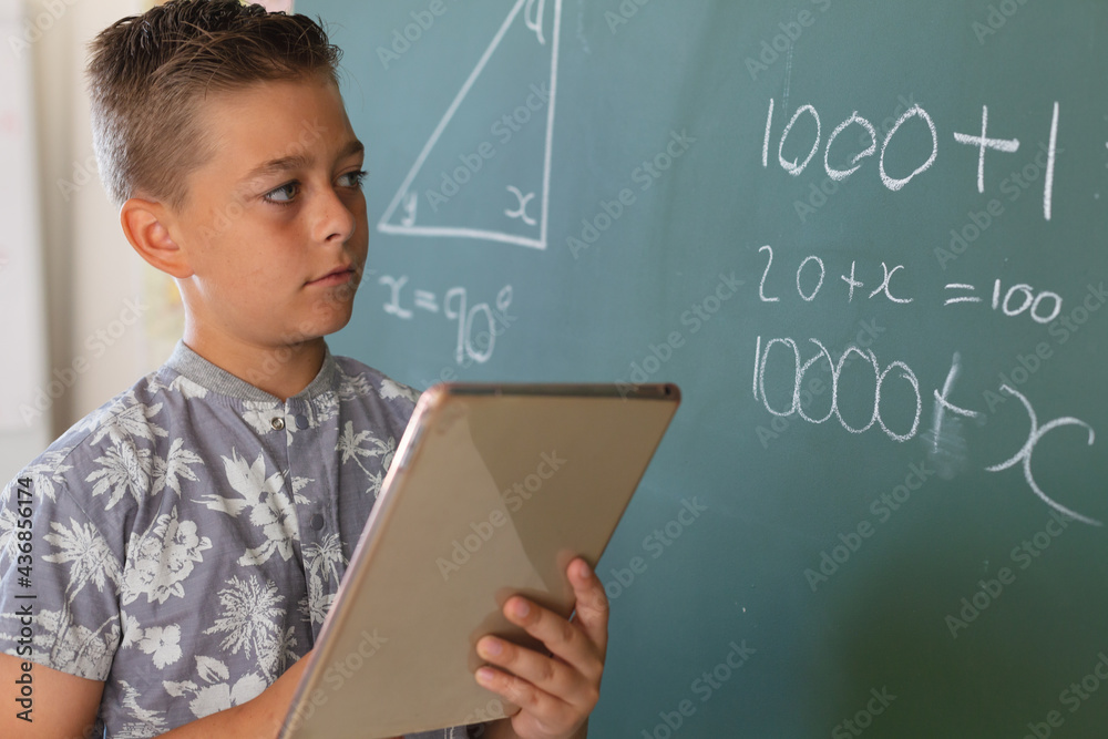 Caucasian boy standing at chalkboard in classroom and using tablet ...