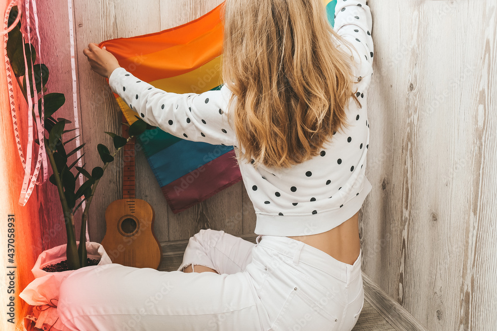 Young blonde millennial hippie woman with rainbow LGBTQ flag at home ...