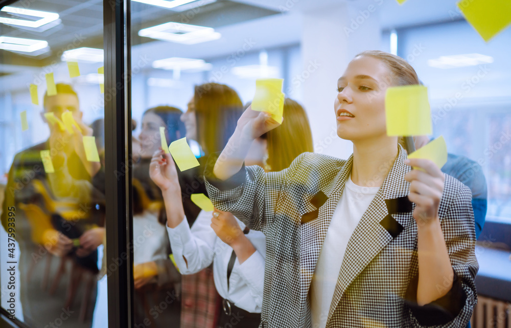 Business colleagues writing on sticky note on glass wall with coworkers ...
