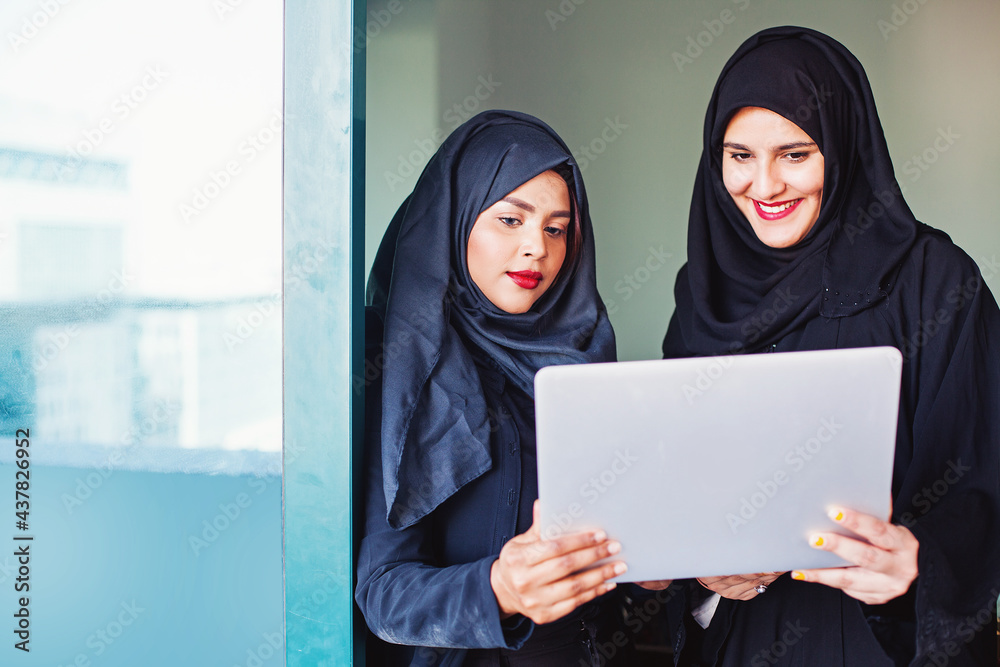 Two Muslim women in modest clothes and hijab holding and using a laptop ...