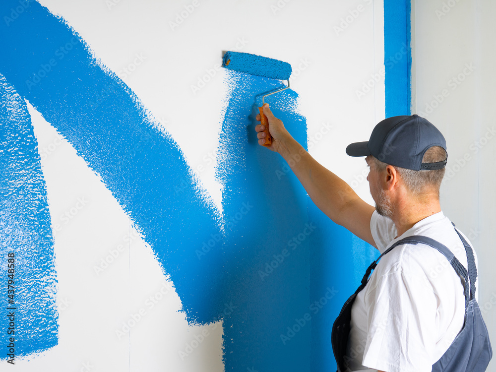 Male painter painting a white wall with a roller in blue color ...