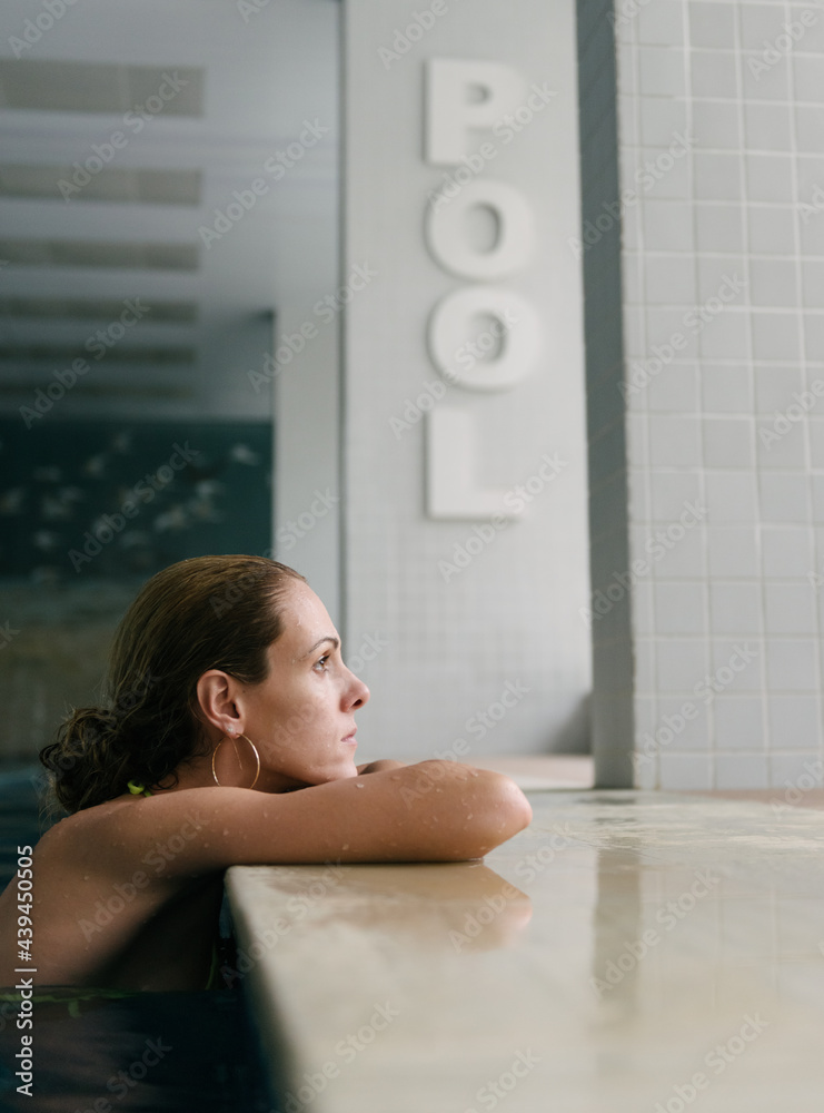 Woman hangs alongside the edge of a n indoor pool – alone, thinking ...
