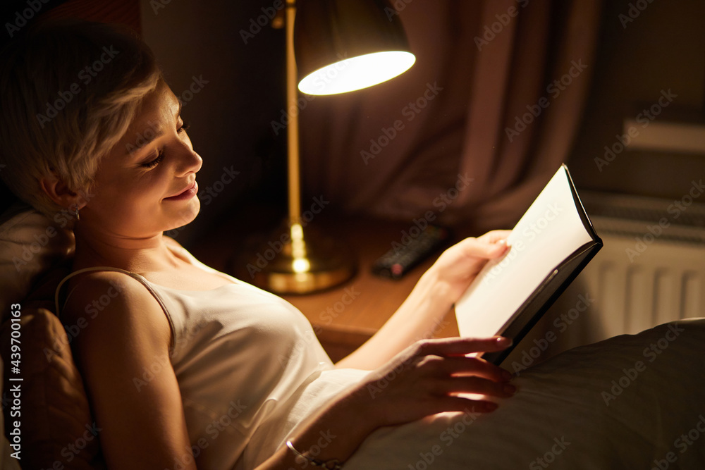 Beautiful female in pajamas lying on bed reading book, alone at night ...