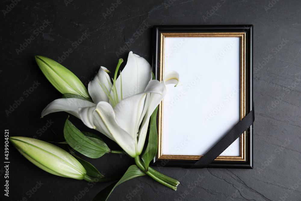 Funeral photo frame with ribbon and white lily on black table, flat lay ...