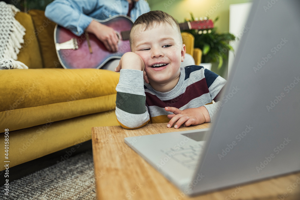 Cute boy watching video on laptop while father playing guitar in living ...