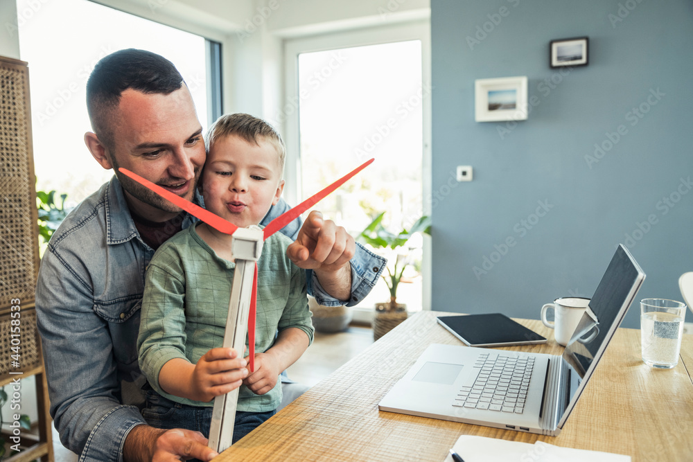 Smiling father sitting with son blowing wind turbine at home Stock ...