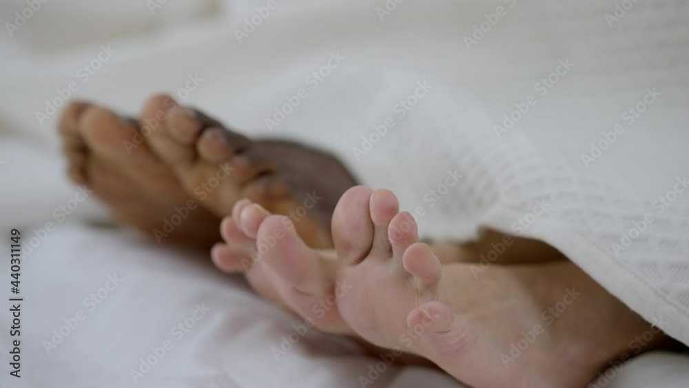 Close-up female Caucasian and male African American feet on bed moving ...