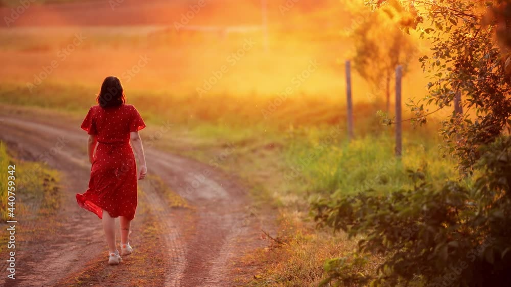 Young Woman in long red dress walking away in the evening sun along a ...